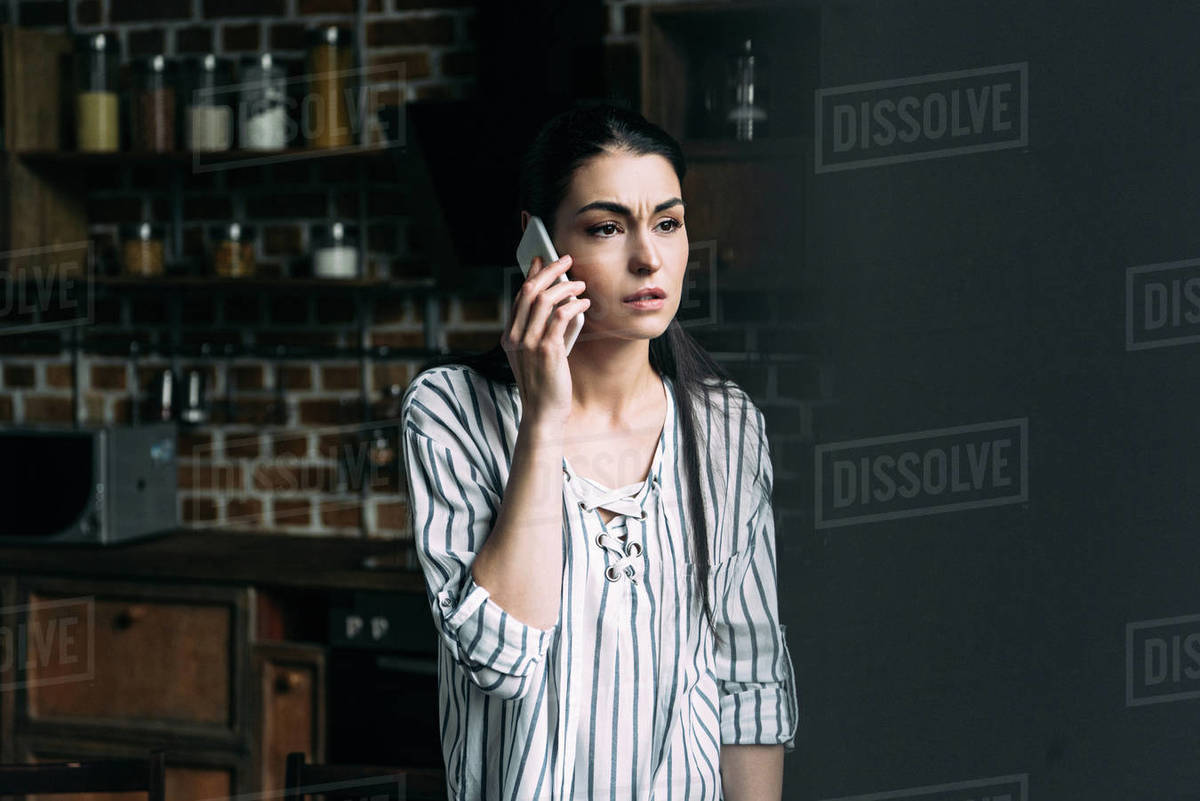 sad young woman talking by phone and looking away on kitchen - Royalty ...