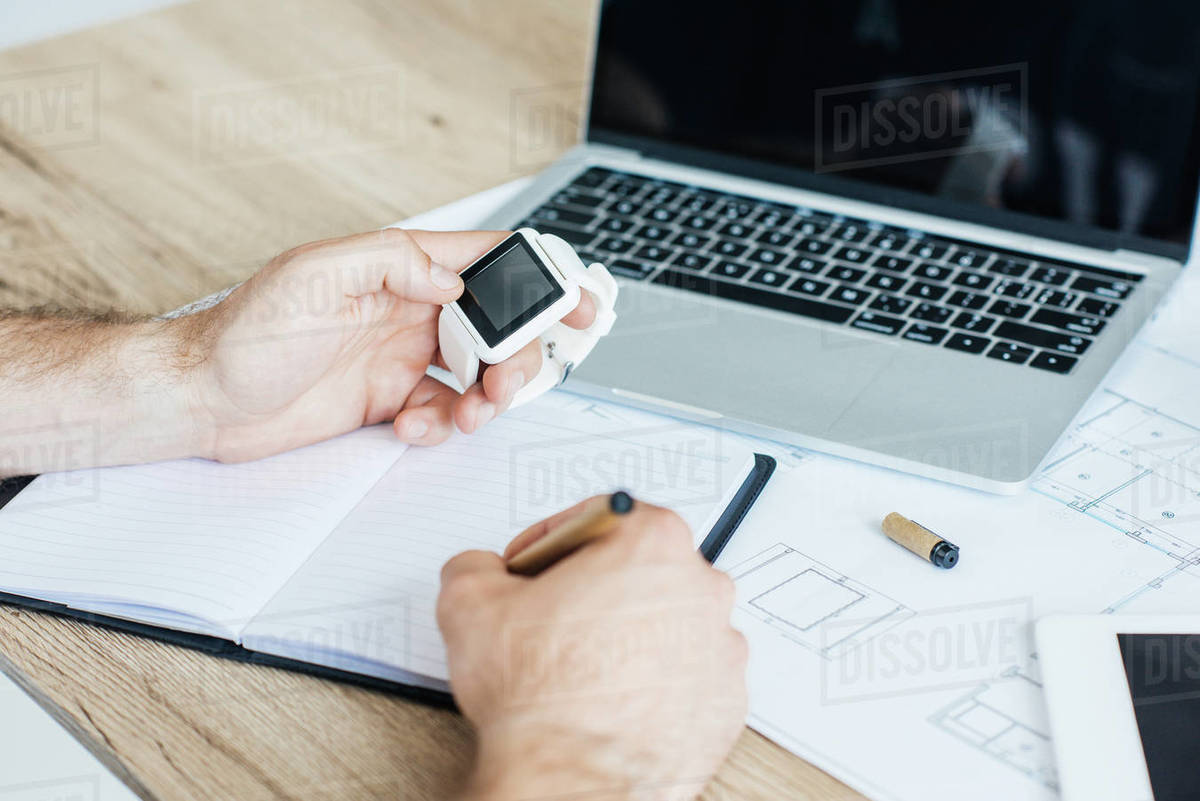 cropped shot of person holding smartwatch and taking notes at workplace ...