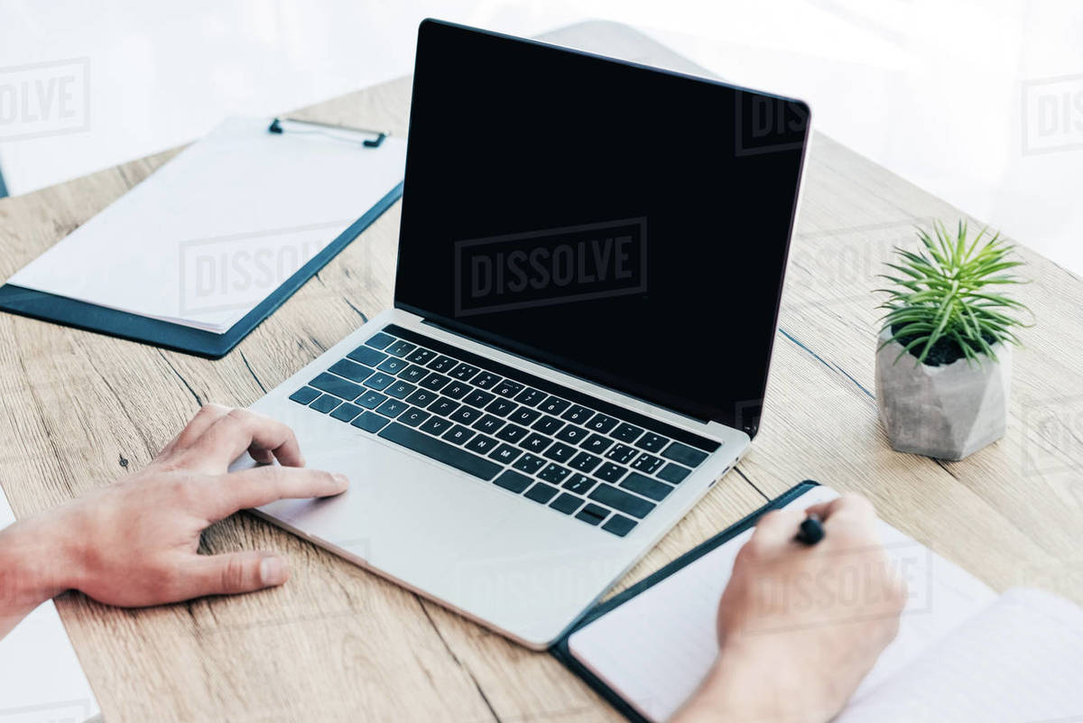 Cropped image of man writing in textbook and using laptop at table with ...