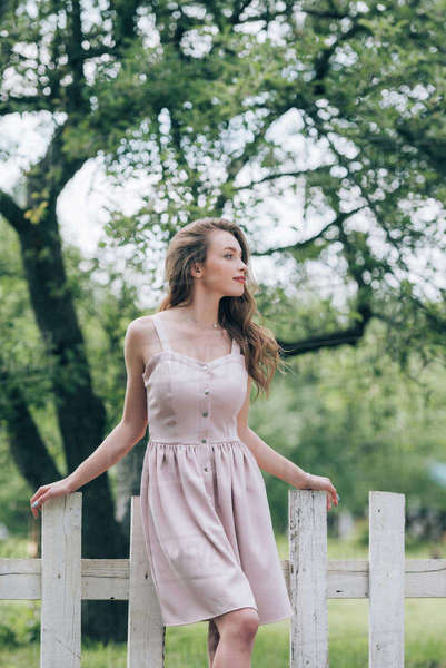 Pensive beautiful woman in stylish dress standing at white fence at ...