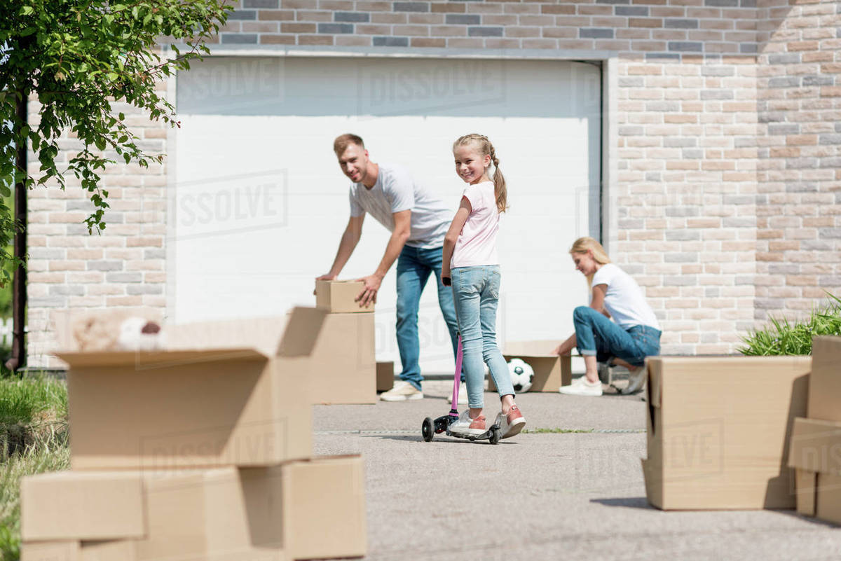 Couple unpacking cardboard boxes while their daughter riding on kick ...