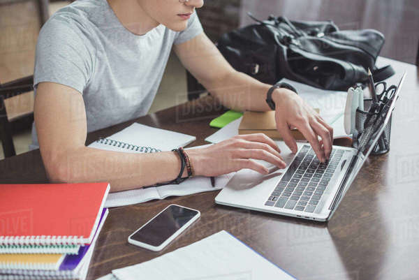 Cropped view of male student doing homework with laptop at table with ...