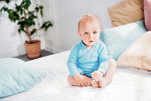 Adorable little baby sitting on bed at home and looking at camera ...