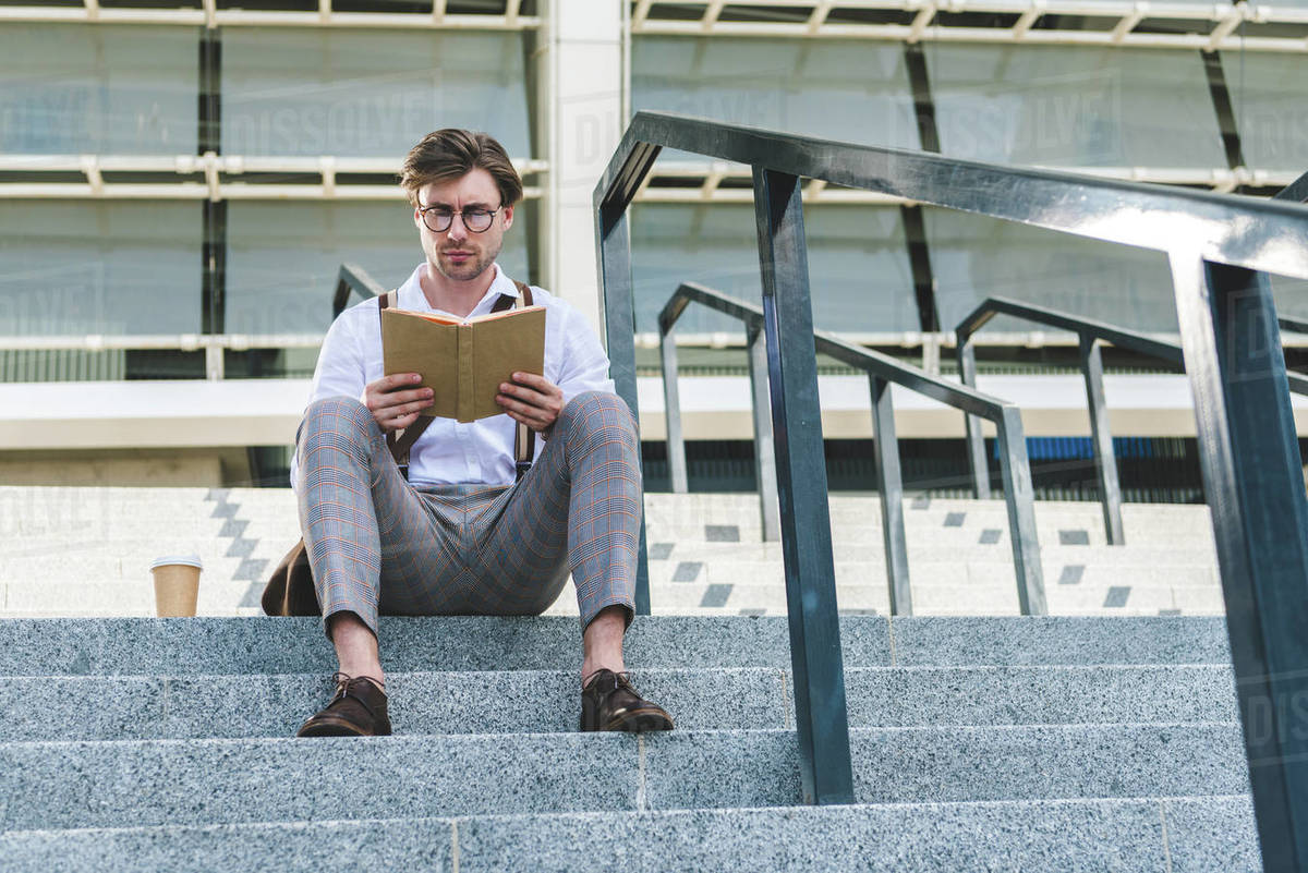 Bottom view of handsome young man with paper cup of coffee reading book ...