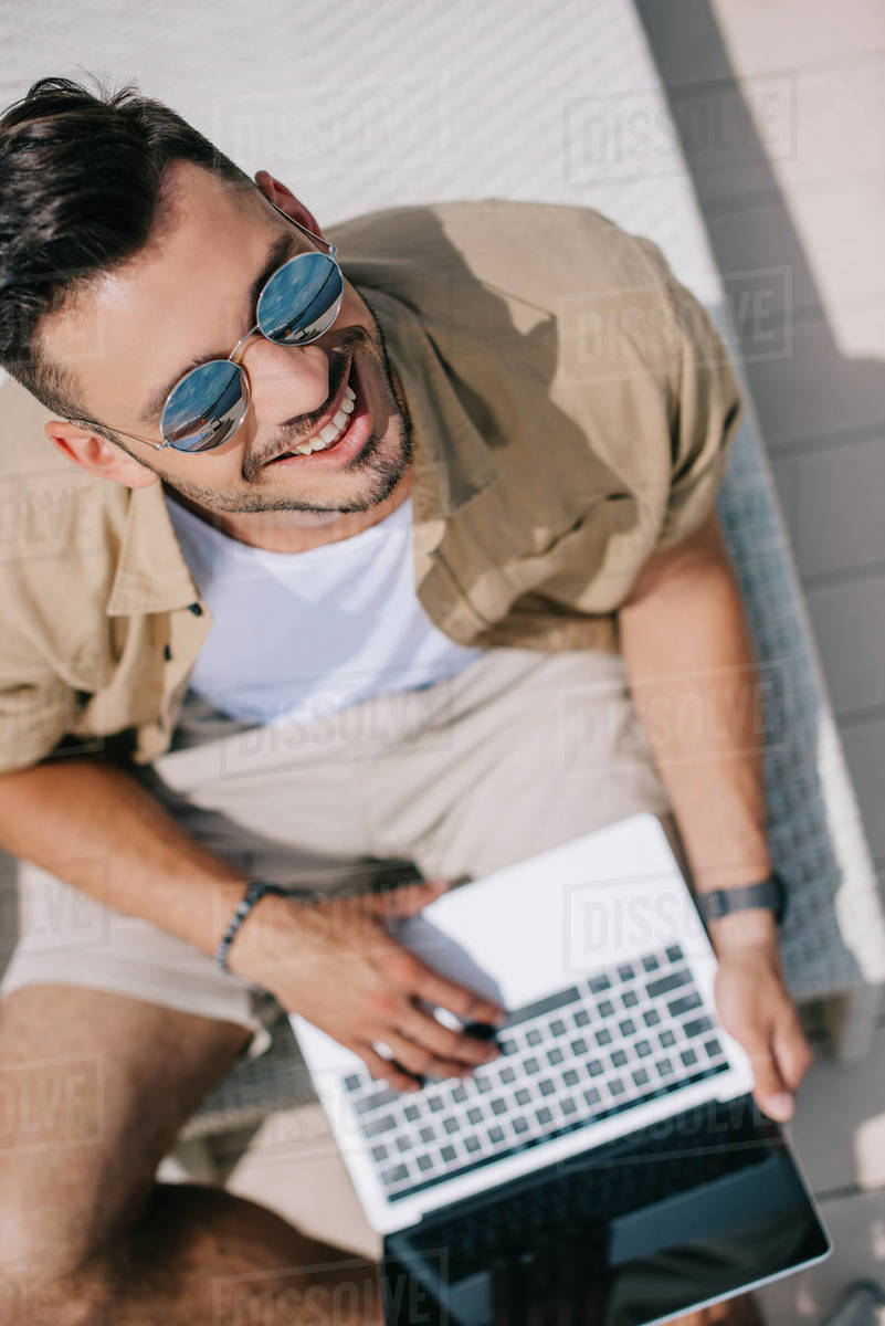 Overhead view of young man in sunglasses using laptop and smiling at ...