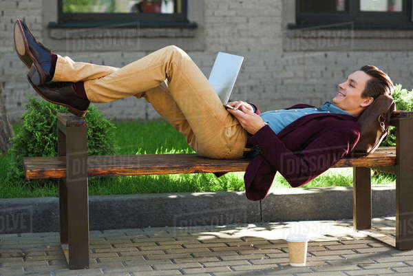 Smiling young freelancer lying on bench and using laptop - Royalty-free ...
