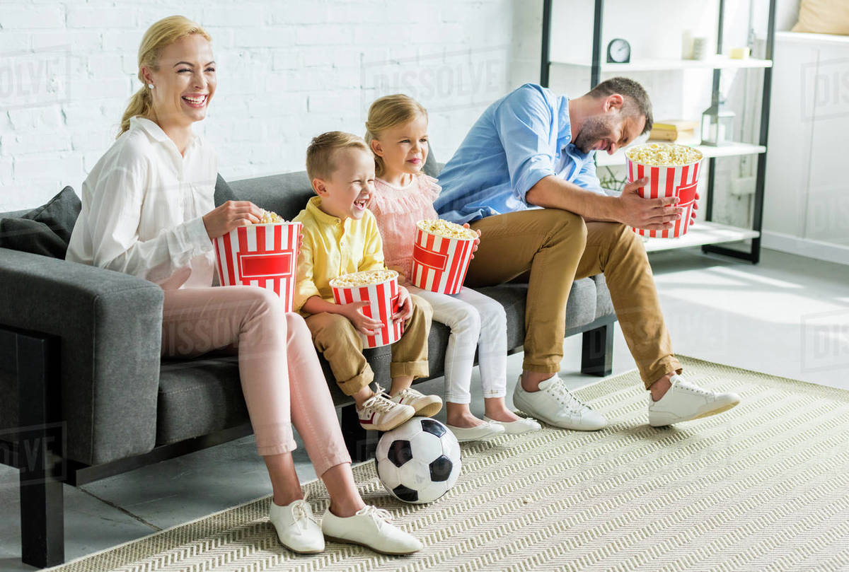 Happy family eating popcorn while sitting together on sofa at home ...