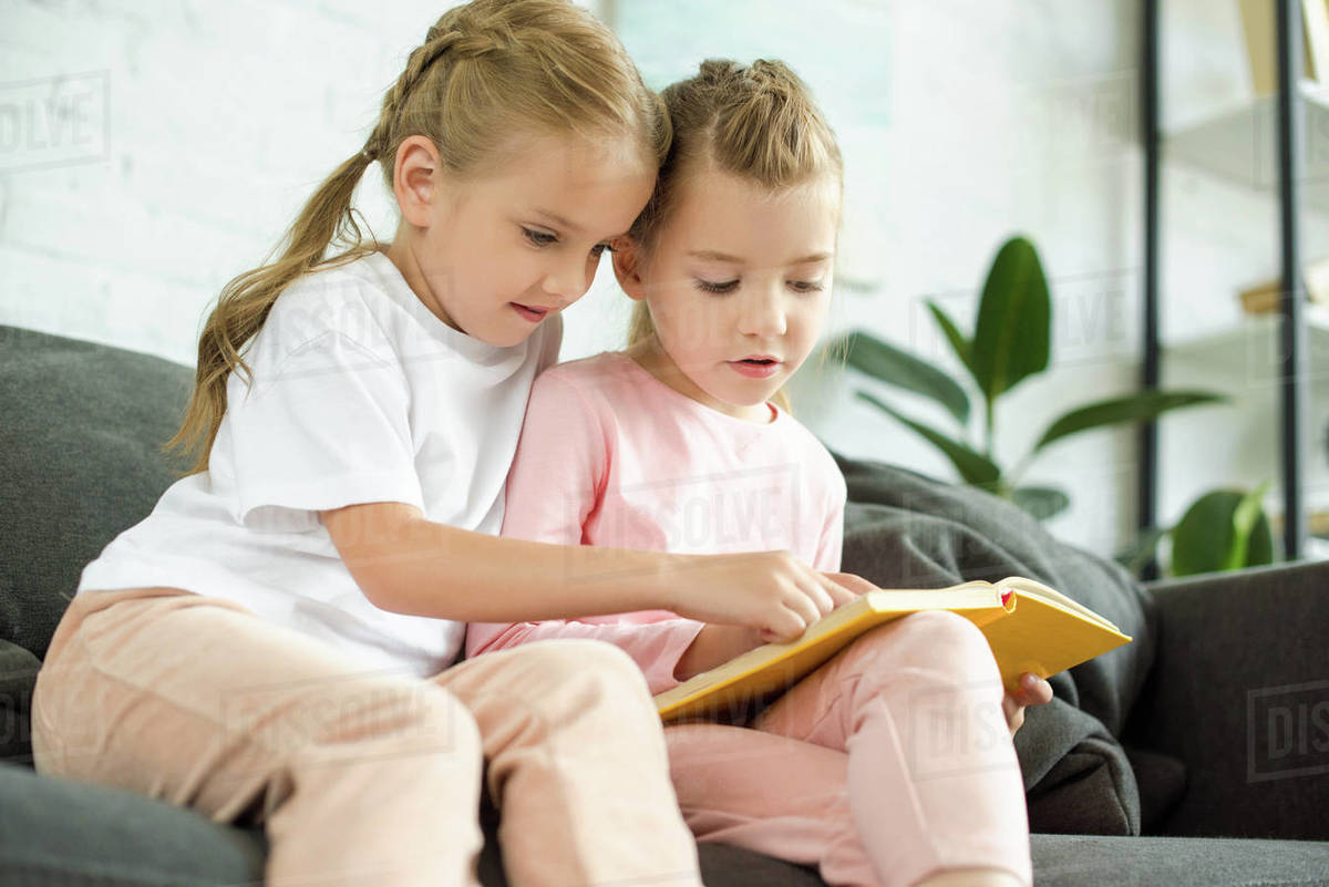 Adorable little sisters reading book together on sofa at home - Royalty ...