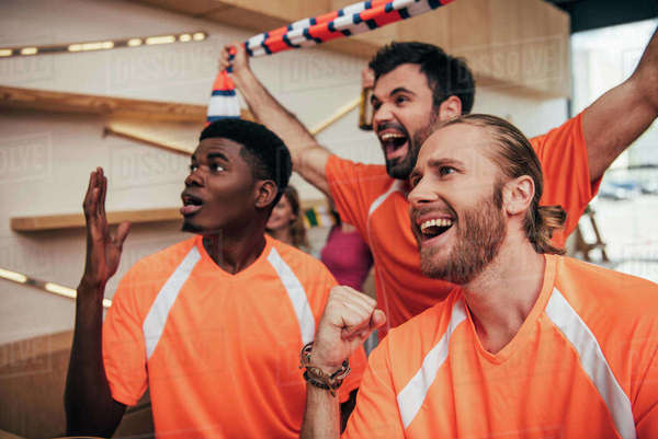 Excited multicultural male football fans in orange t-shirts celebrating ...