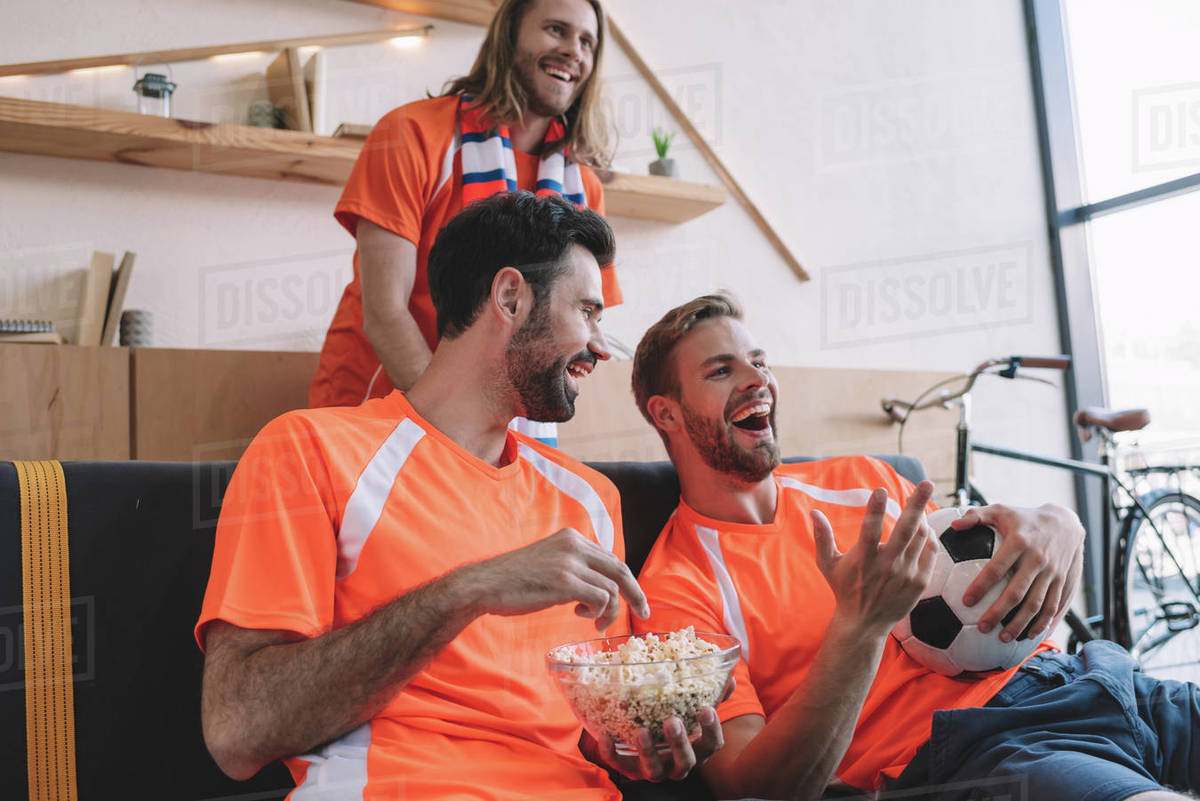 Smiling male football fans in orange t-shirts with ball and popcorn ...