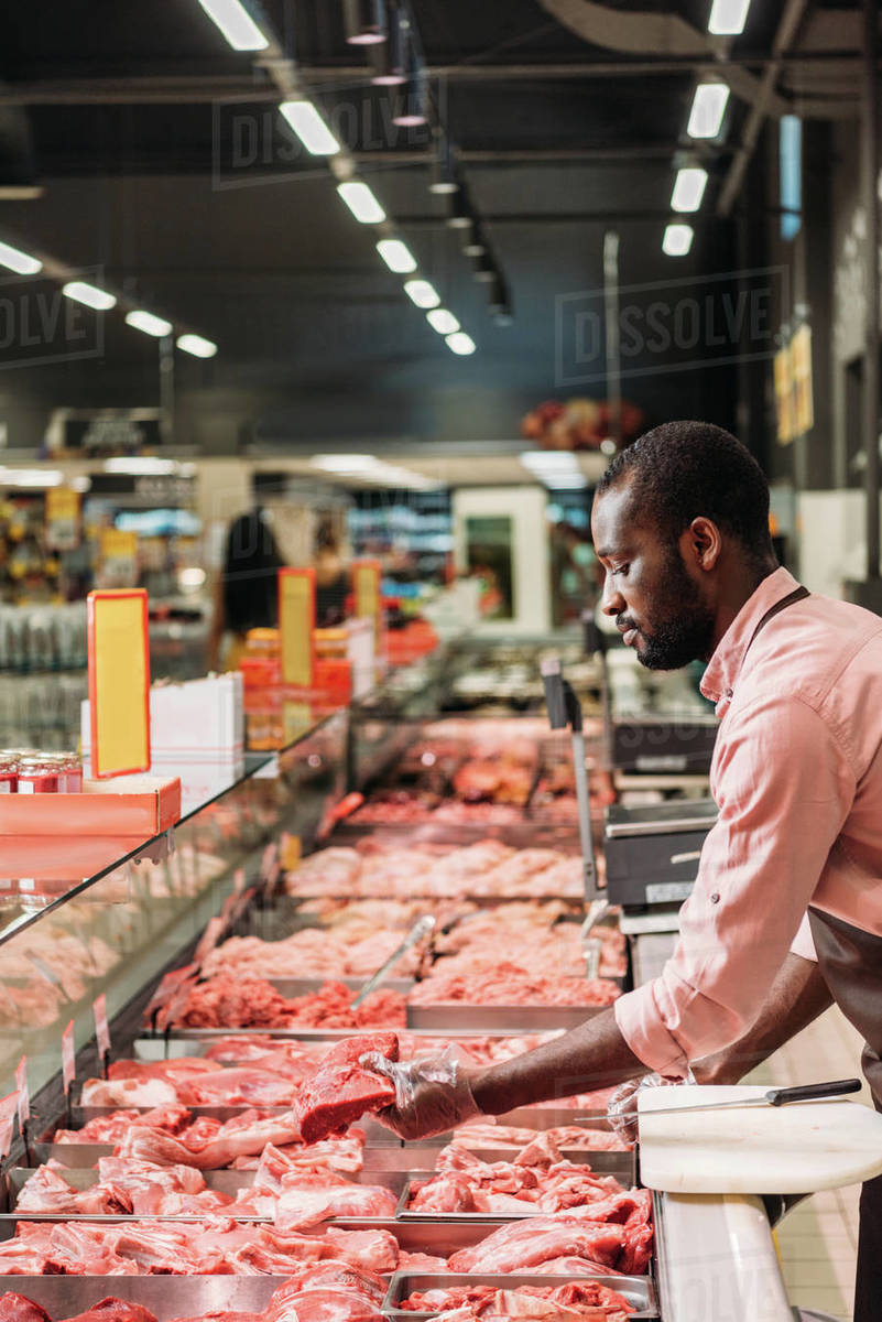 Focused african american male butcher in apron taking steak of raw meat ...