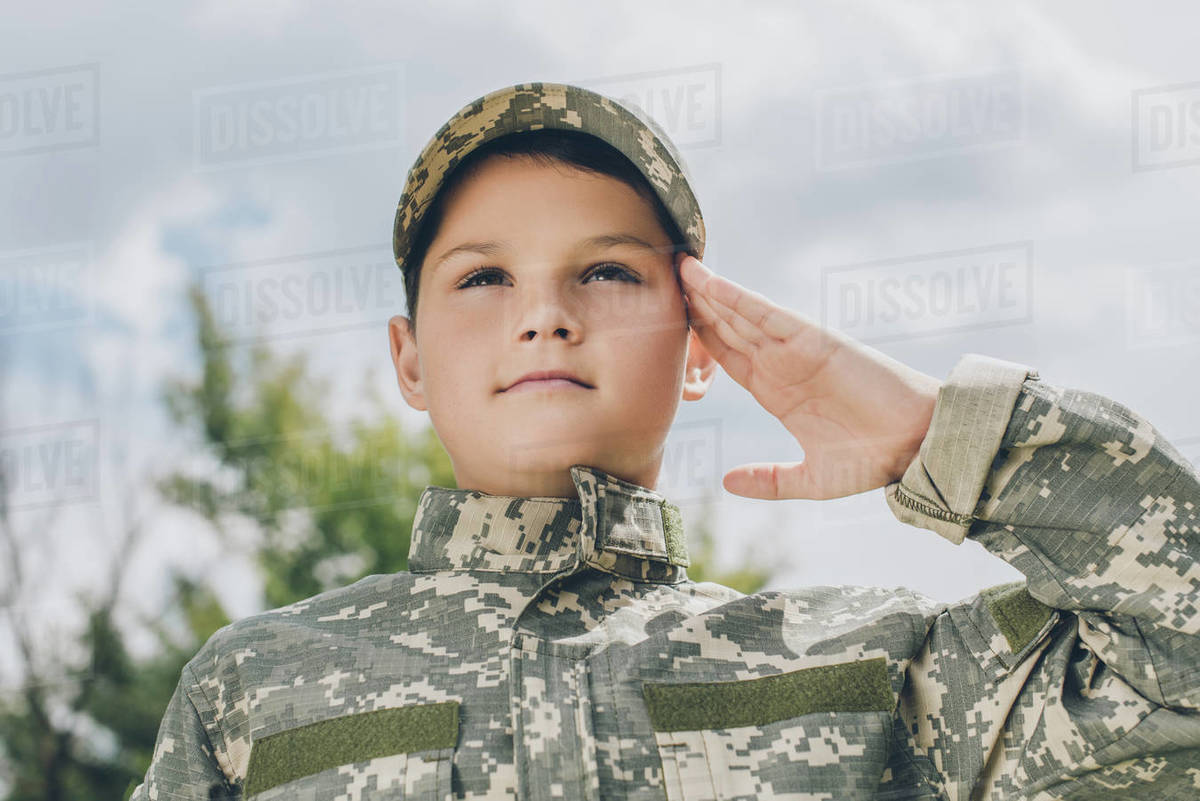 portrait of little boy in camouflage clothing saluting with cloudy sky ...