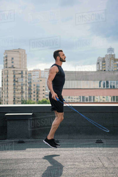 Side view of handsome sportsman training with jumping rope on roof ...