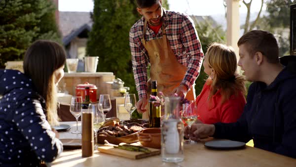 Handsome man brings a plate with a food on a table full of happy people ...