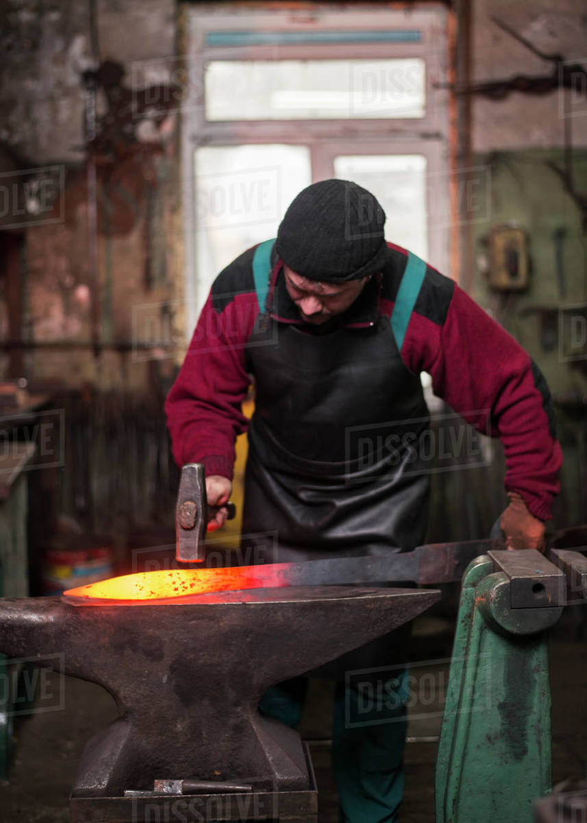 Blacksmith working with a hammer with hot iron piece making a sword ...