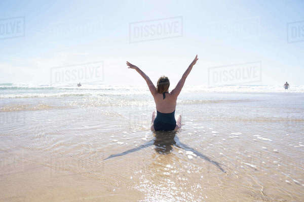 Rear view of a woman sitting on beach in shallow water with her arms in ...