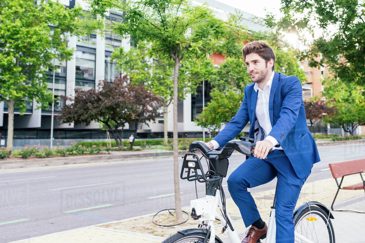 Young smiling man in elegant suit riding electric bicycle on pavement ...