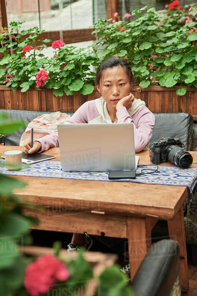 Concentrated Asian female journalist sitting at table with digital ...