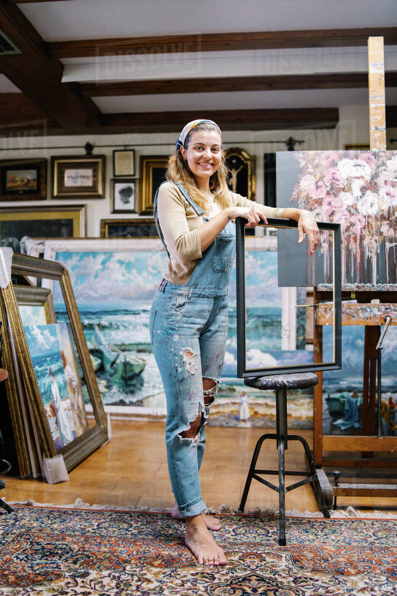 Full length of creative young female artist in denim overall holding ...