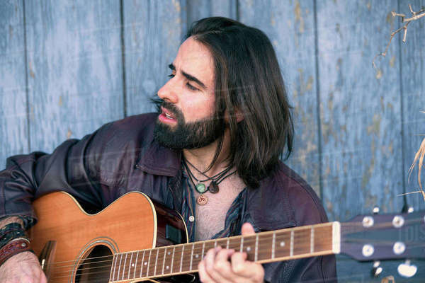 Bearded guitarist with long hair sitting next to shabby wall holding ...