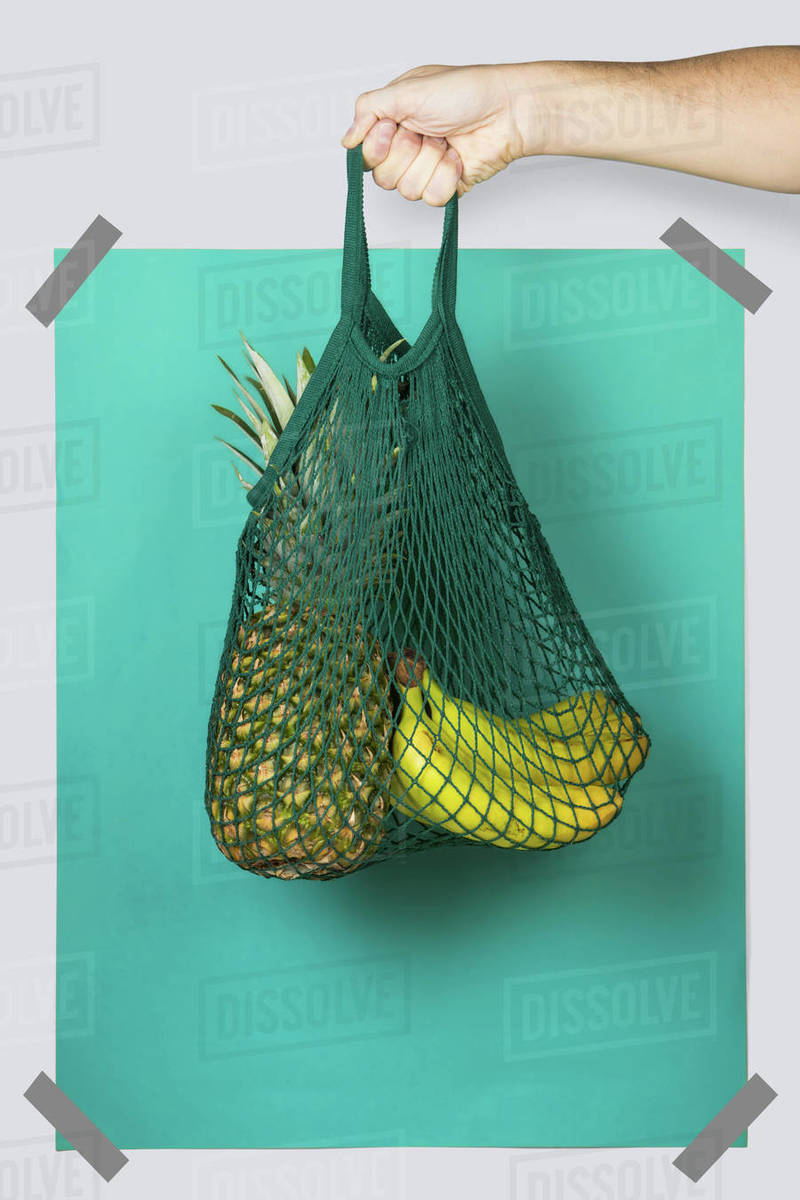 Unrecognizable person carrying net bag with ripe pineapple and bananas ...