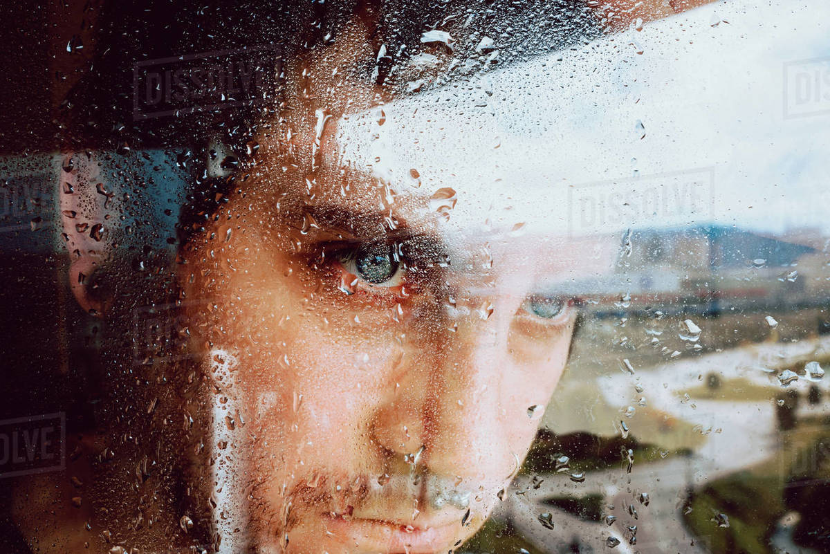 Closeup of sad desperate young man looking away through wet window ...