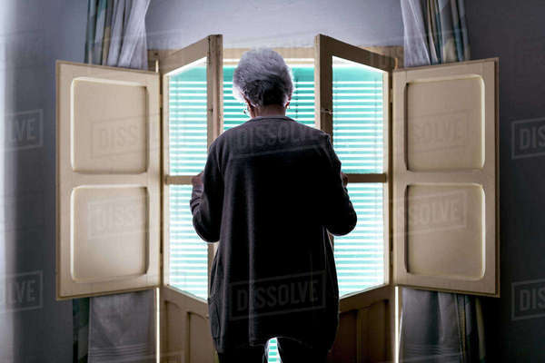 Back view of unrecognizable senior female opening window with shutters ...