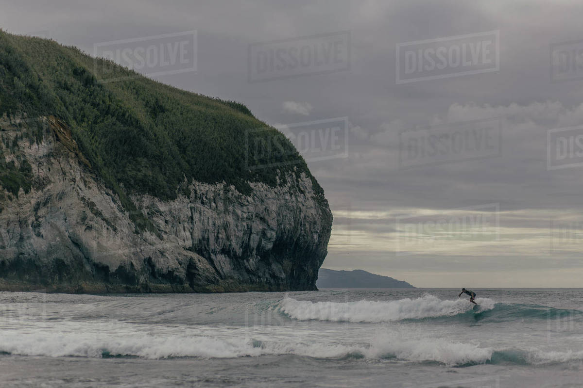 Person surfing on high waves in rough sea near huge rock on background ...