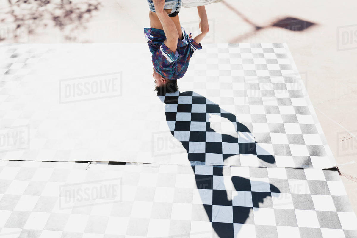 From above shadow of crop young guy doing headstand on checkered floor ...