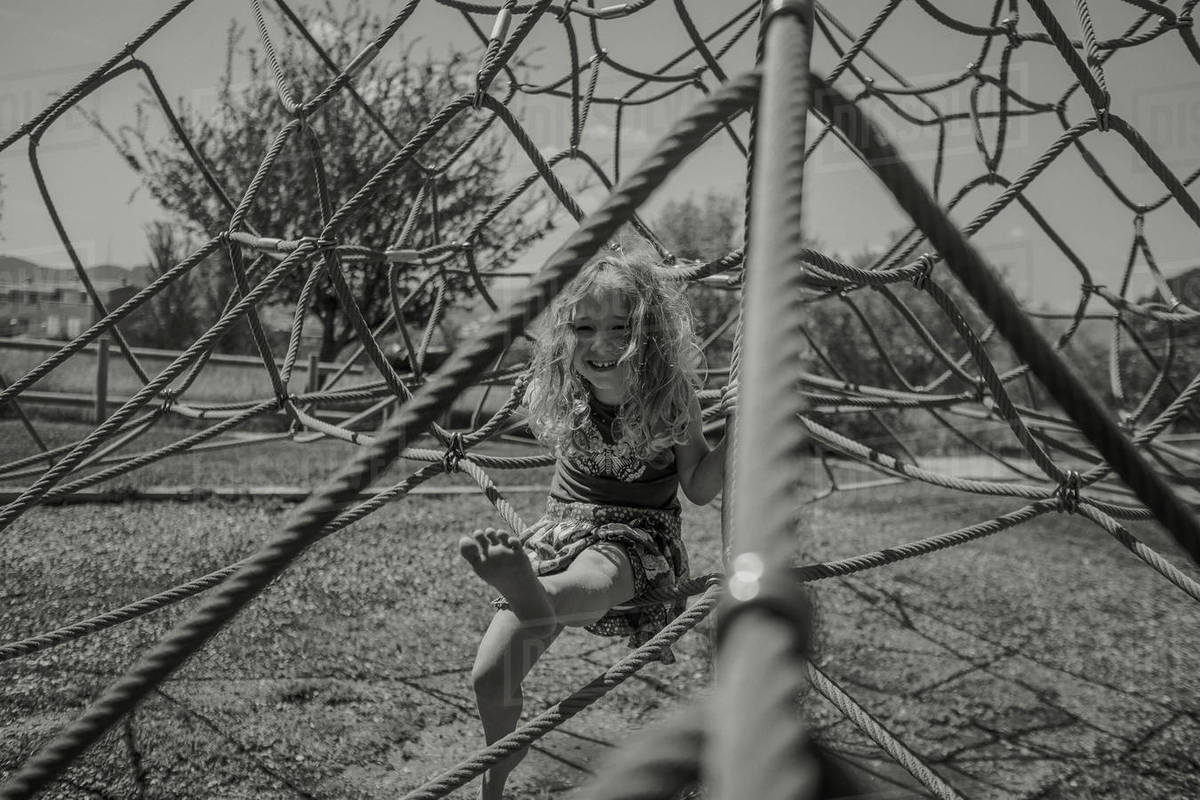 Curly enthusiastic barefoot girl sitting on rope of playground ...