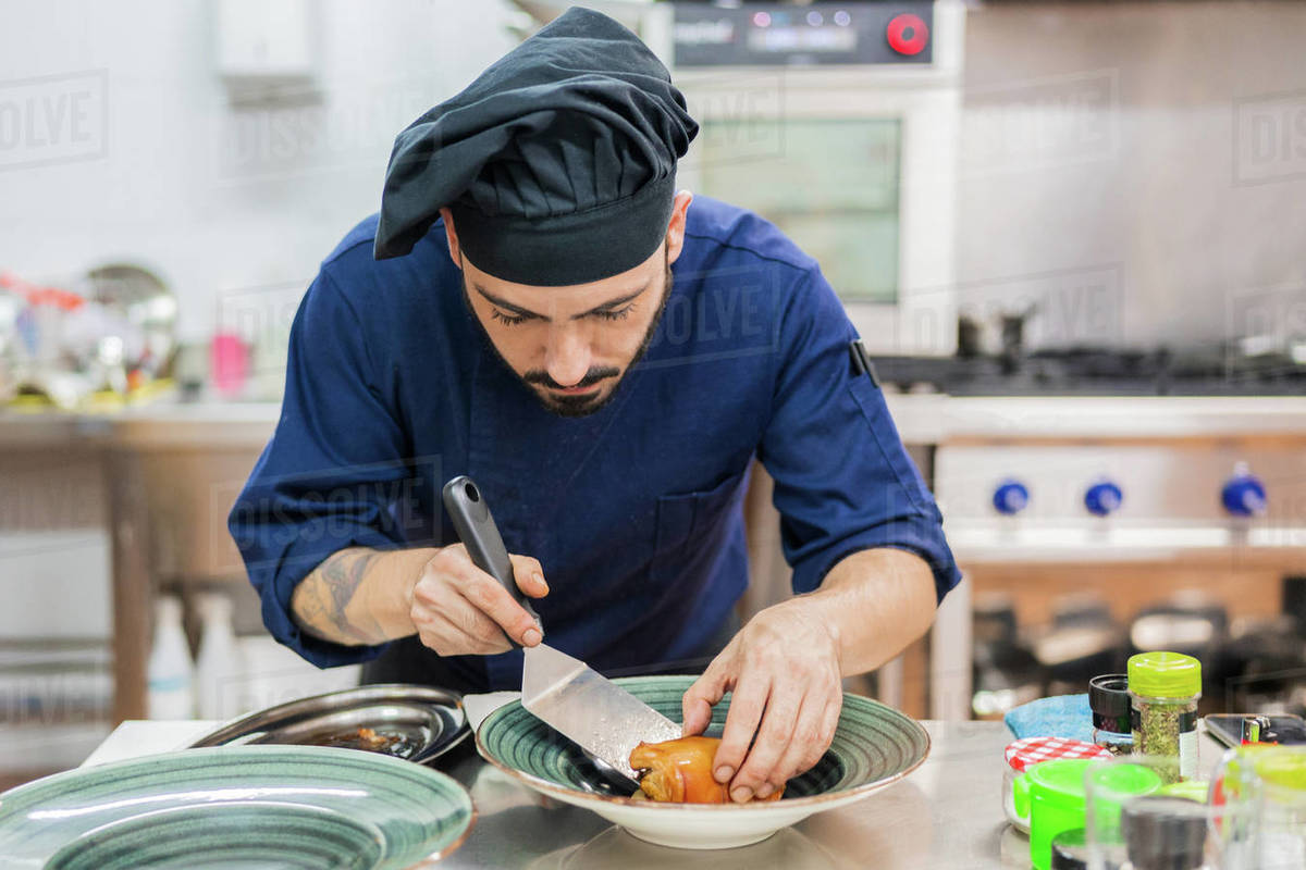 Focused professional male cook using spatula and putting food on plate ...