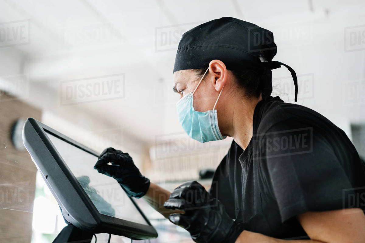 Side view serious female cashier wearing gloves and mask tapping on ...