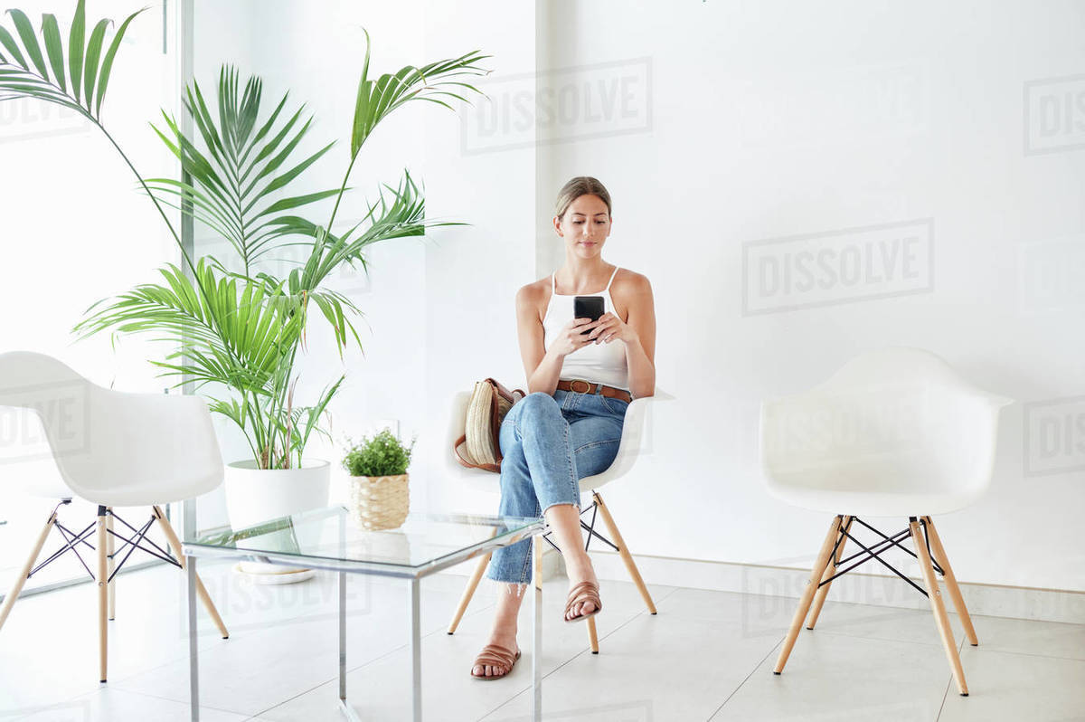 Full body female client sitting on chair and browsing on smartphone ...