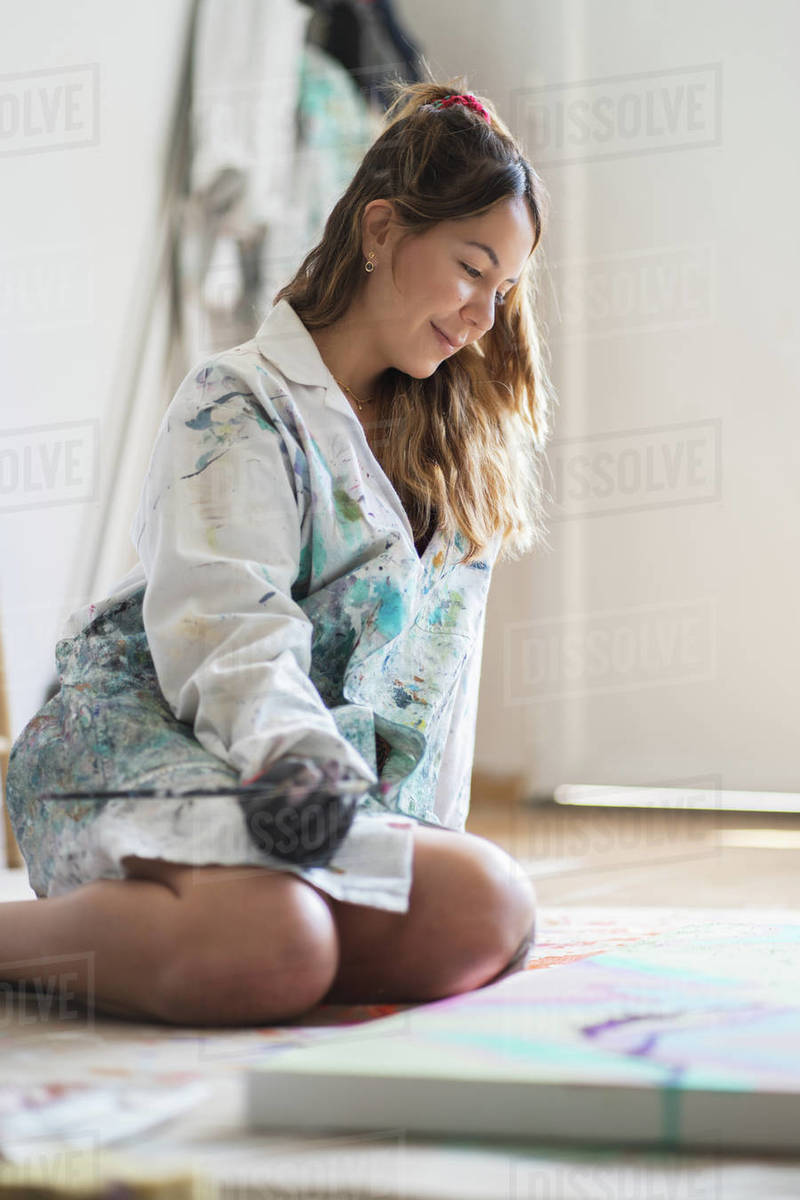 Cheerful barefoot female artist in dirty robe and gloves lying on ...