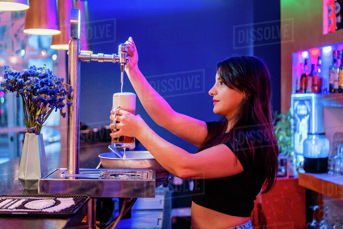 Side view of young female barkeeper pouring beer into glass while ...