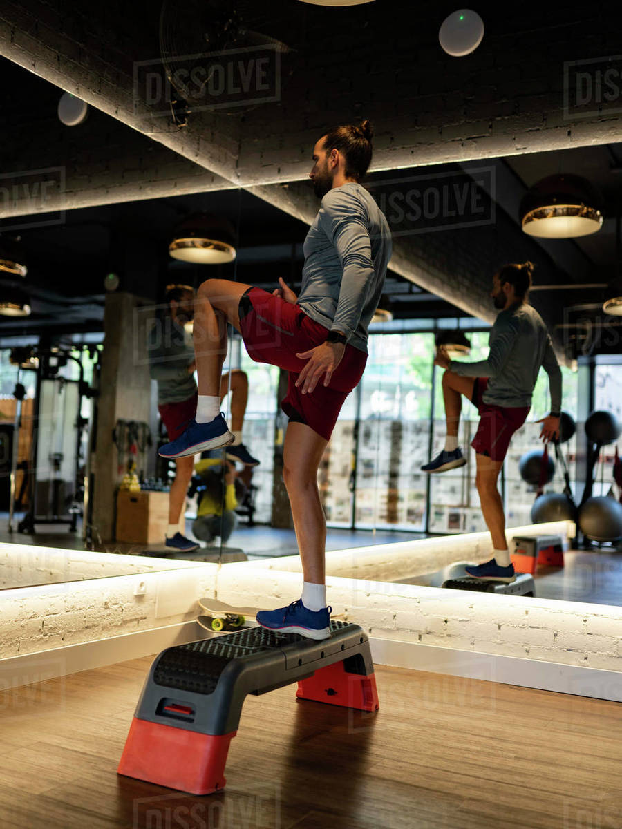 Full body male athlete standing on step and lifting leg near mirror ...