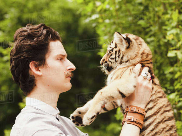 A man with a mustache looks into the face of a tiger cub outdoors ...