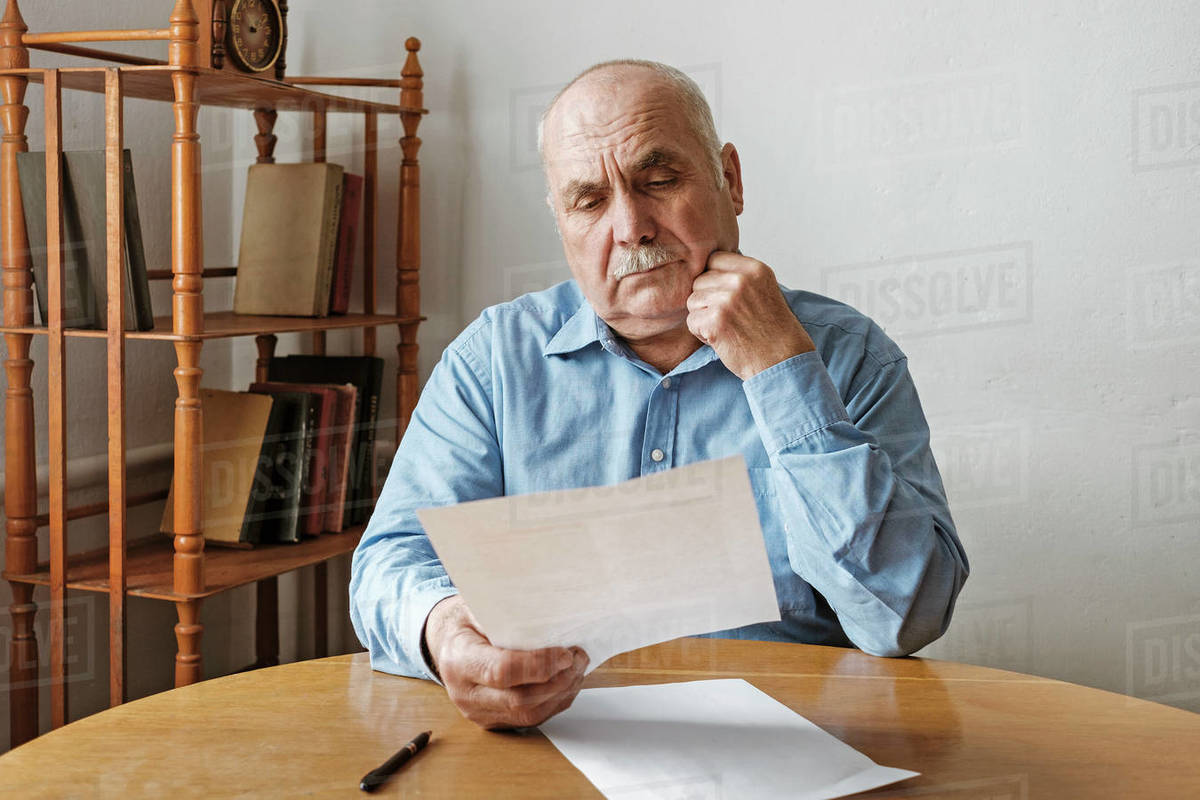 Thoughtful old man reading a handheld paper document with a look of ...