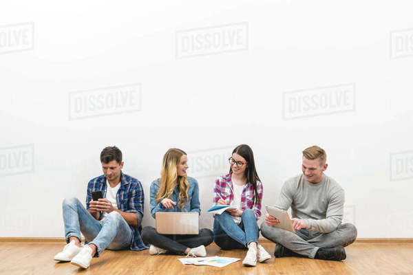 The happy friends with gadgets sit on the floor on the white wall ...