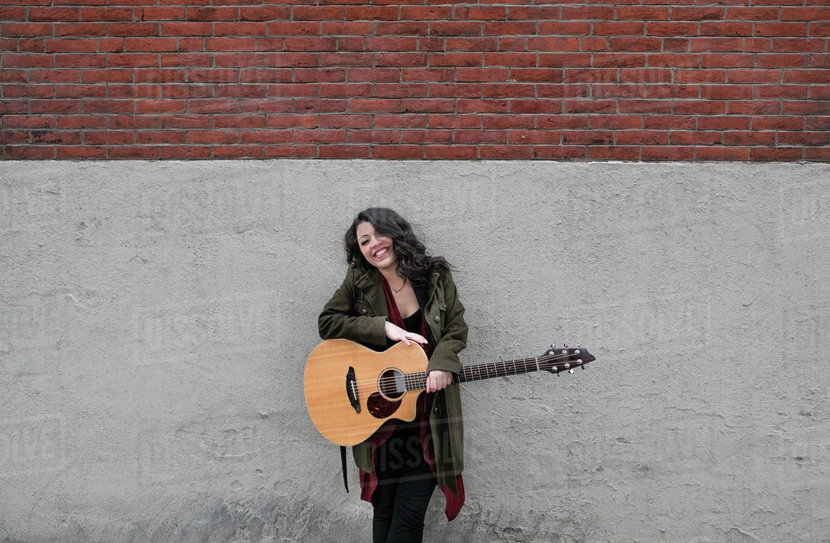 Portrait of young woman with acoustic guitar leaning against wall ...