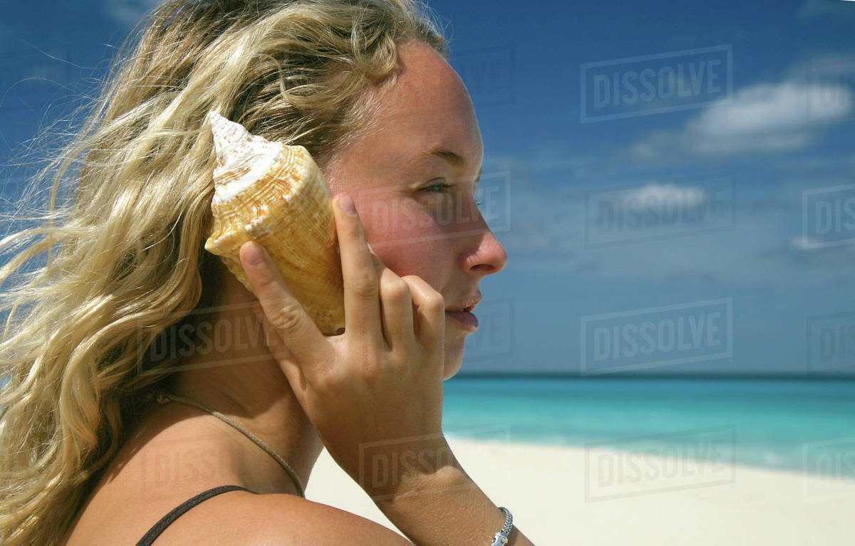 Woman holding shell by ear at beach - Stock Photo - Dissolve