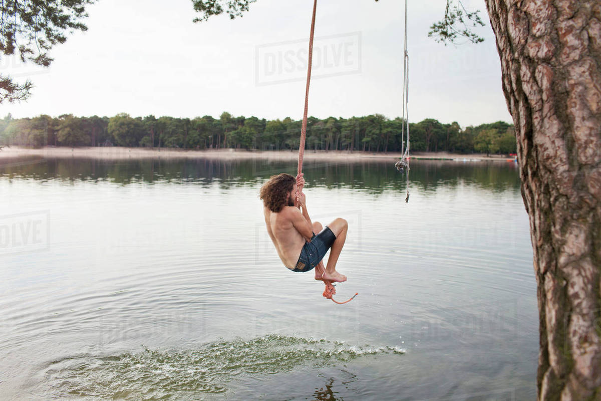 Young man rope swinging above lake - Stock Photo - Dissolve