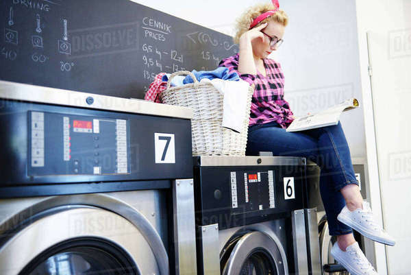Woman sitting on top of washing machine reading magazine at laundrette ...