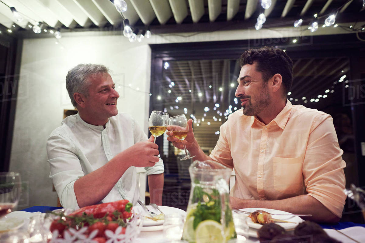 Two men sitting at dinner table, holding wine glasses, making toast ...