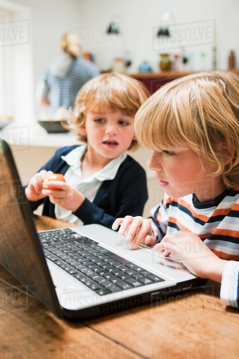 Young boy using laptop with his younger brother looking across at the ...