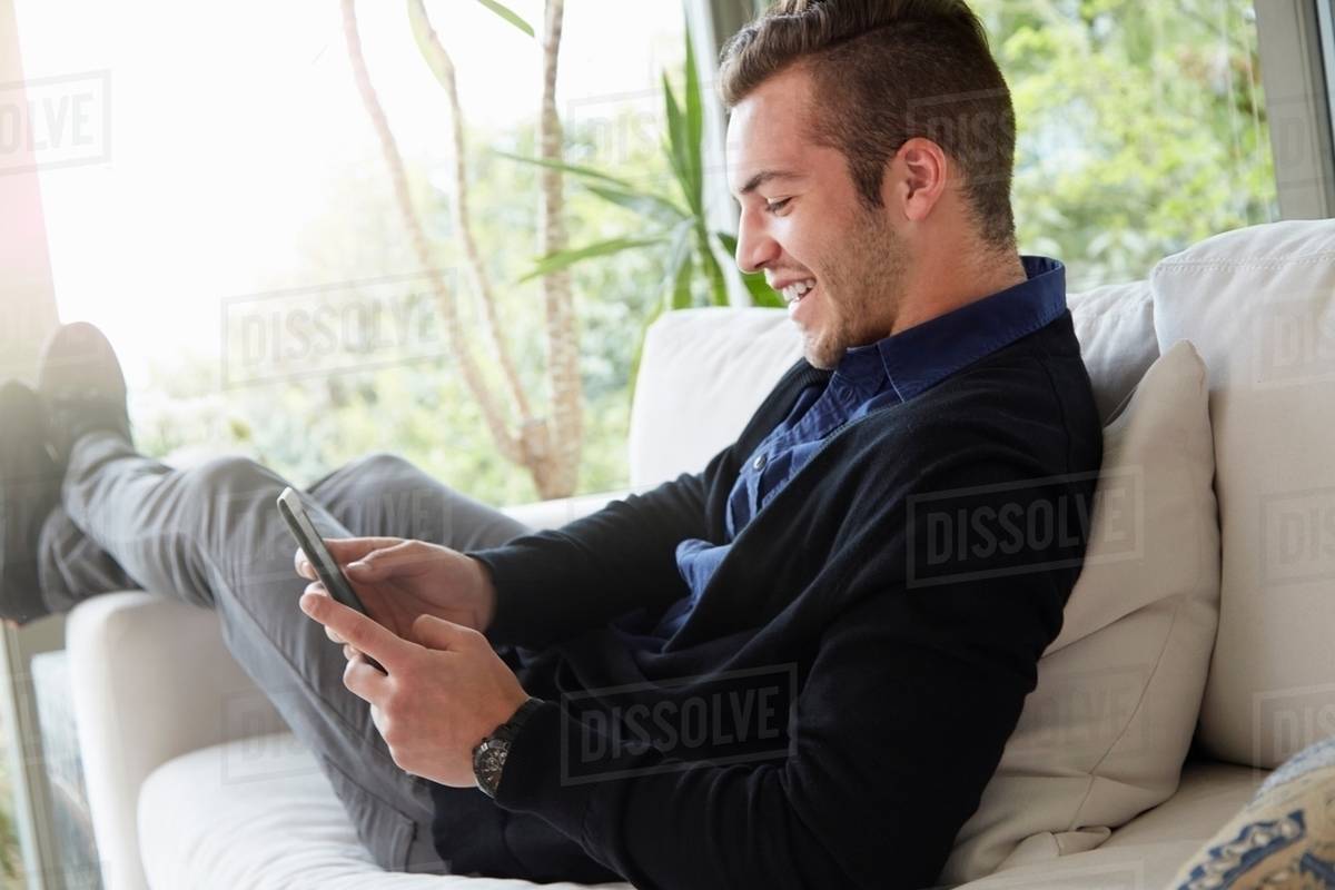 Portrait of man relaxing on sofa with feet up looking at digital ...