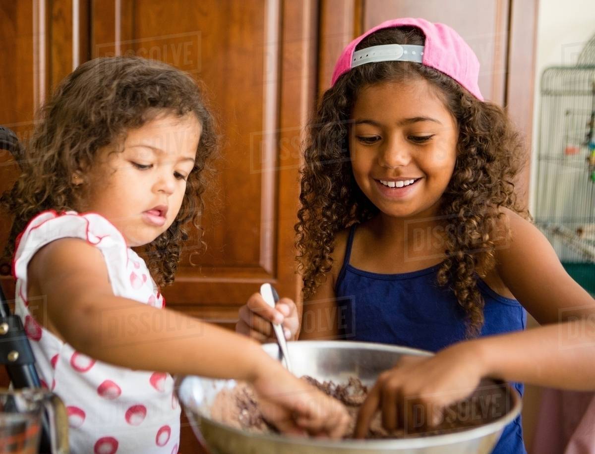 Sisters dipping fingers into chocolate cake mix looking down smiling ...