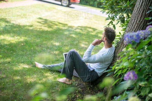 Young man sitting against tree, using laptop - Royalty-free Stock Photo ...