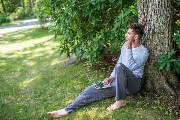 Young man sitting against tree, using laptop - Royalty-free Stock Photo ...
