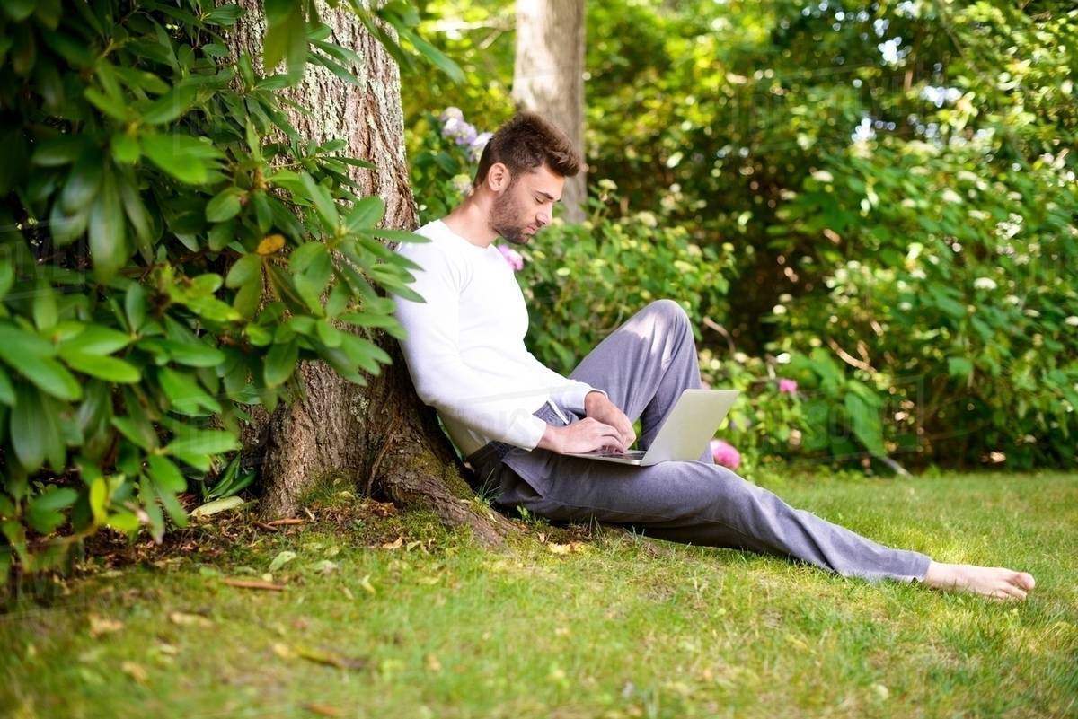 Young man sitting against tree, using laptop - Royalty-free Stock Photo ...
